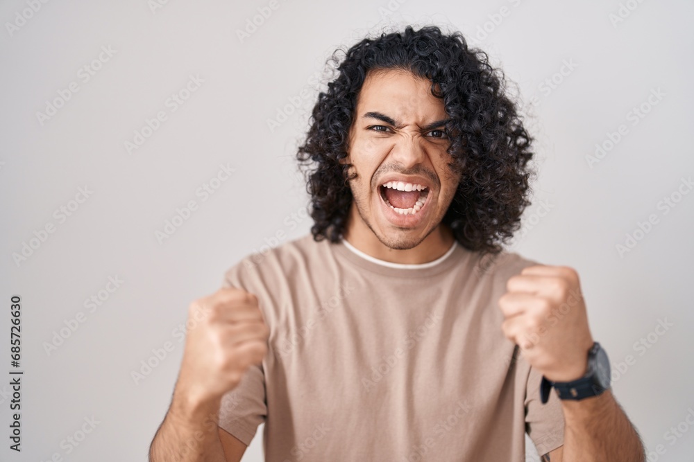 Hispanic man with curly hair standing over white background angry and ...