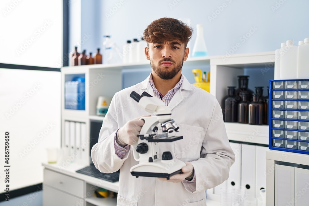 Arab man with beard working at scientist laboratory holding microscope skeptic and nervous ...