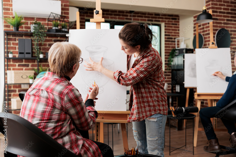 Female Art Teacher Standing Near Easel Explaining Senior Woman How To Draw Still Life Objects