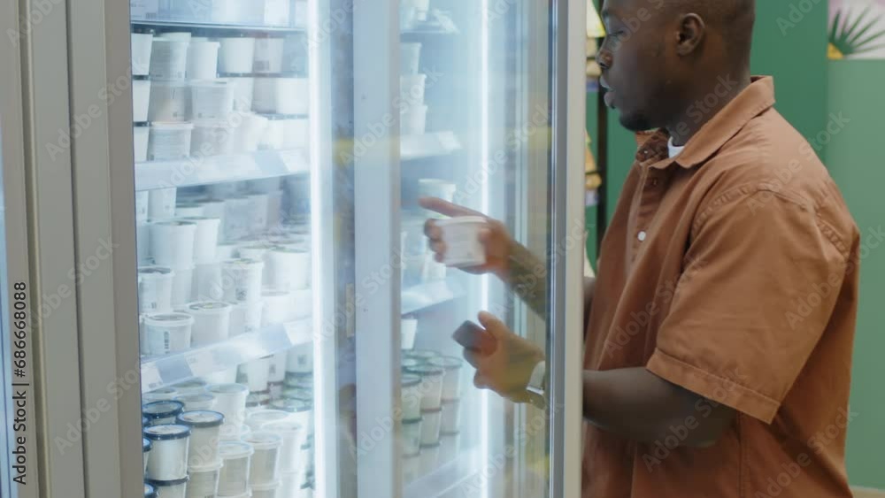 Medium shot of young Black man opening supermarket fridge and taking ...