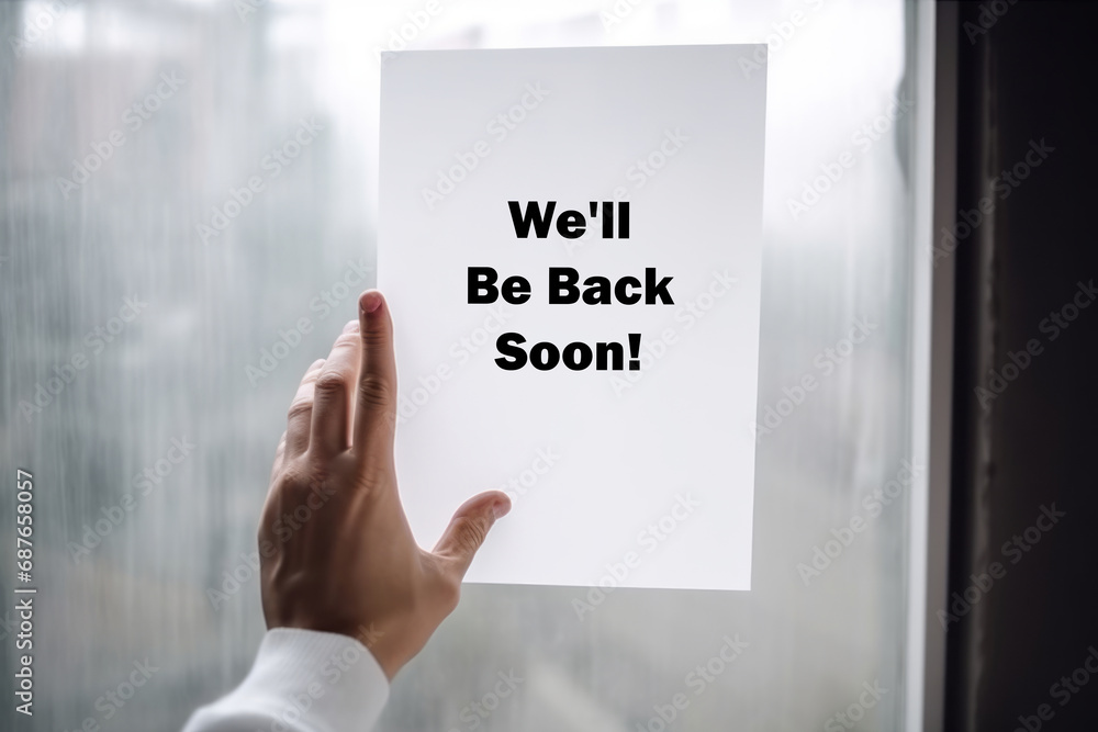 Hands posting “We'll Be Back Soon!” sign on glass door or window. A ...