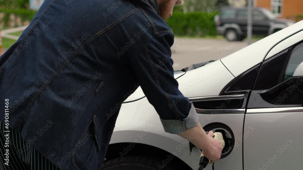 Bearded man unplug power connector from an electric car. Guy in denim ...