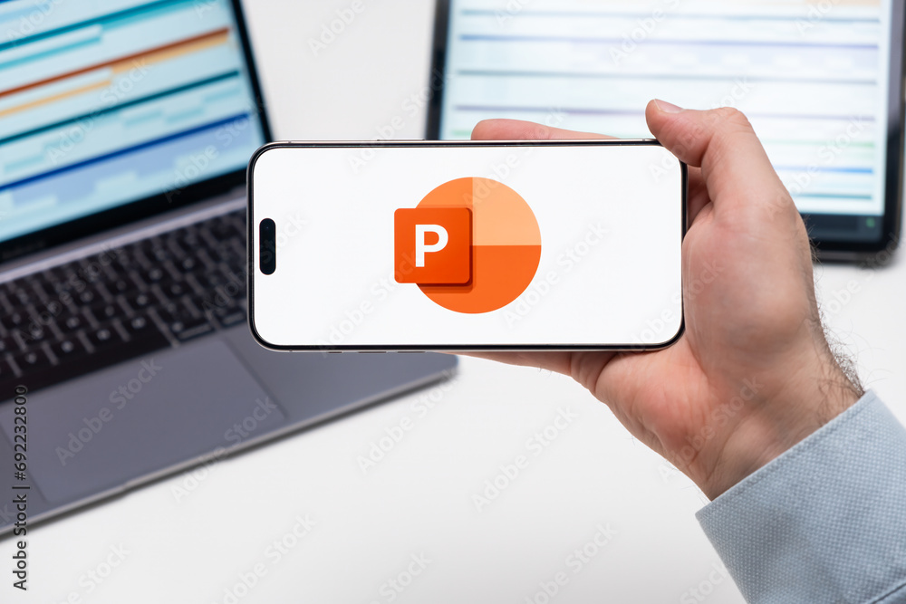 Powerpoint logo on the screen of a smartphone in mans hand with laptop ...