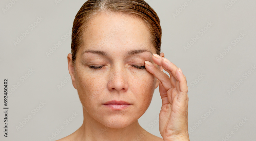 Middle aged caucasian woman of 40s with closed eyes holding finger on eyelid to show eyes aging ...
