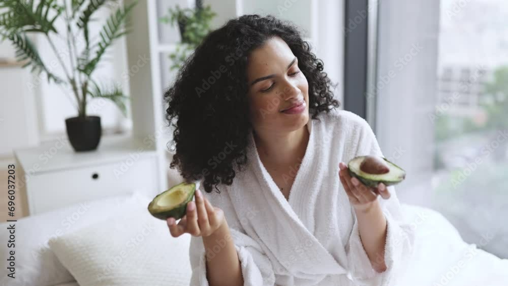Beautiful calm multiracial female in robe sniffs avocado halves in hand ...