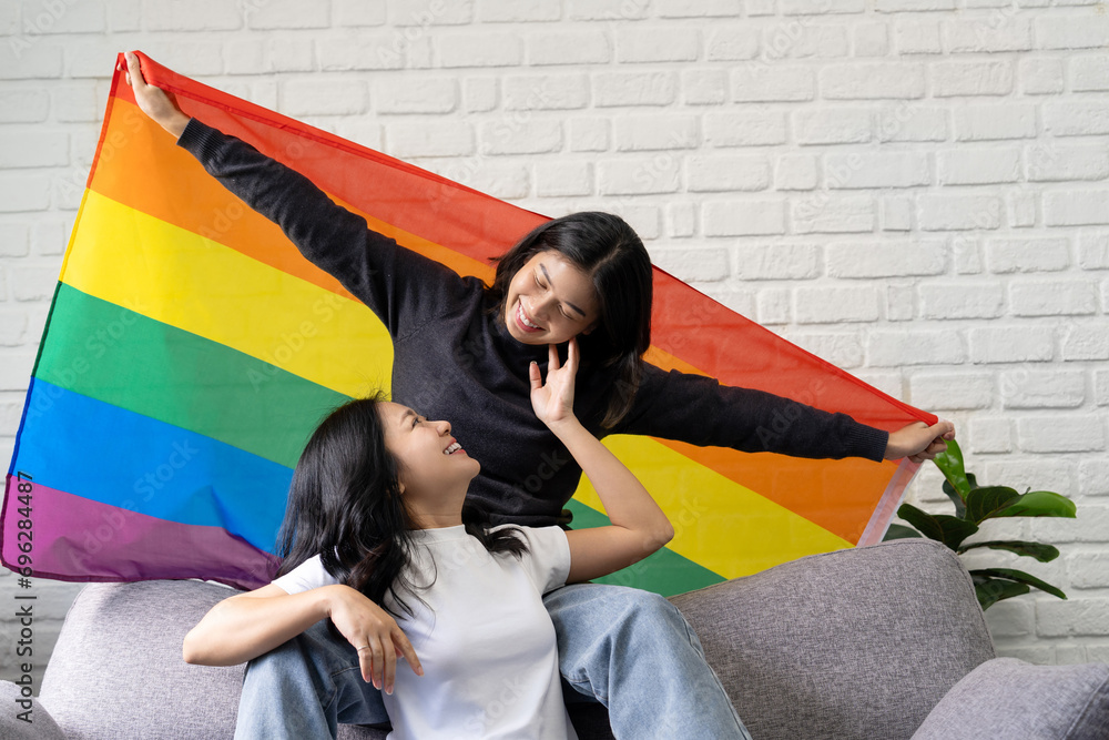 Happy Asian LGBTQ couple sitting on sofa and waving rainbow LGBT Pride ...