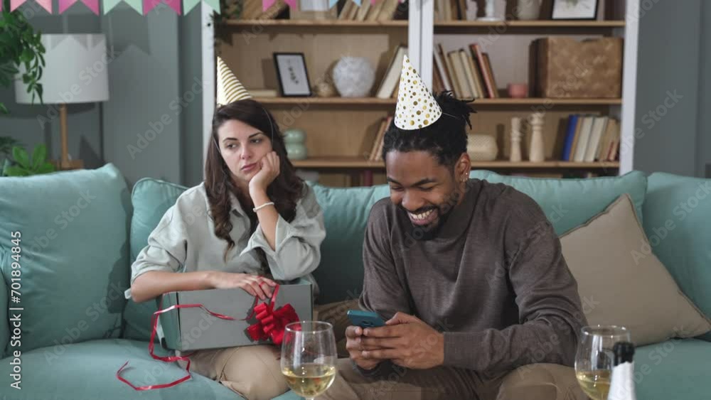 Two young people sitting on sofa with birthday caps, sad because no one ...