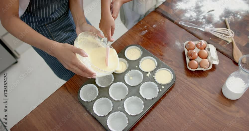 Overhead of biracial mother and daughter pouring cake mix into forms ...