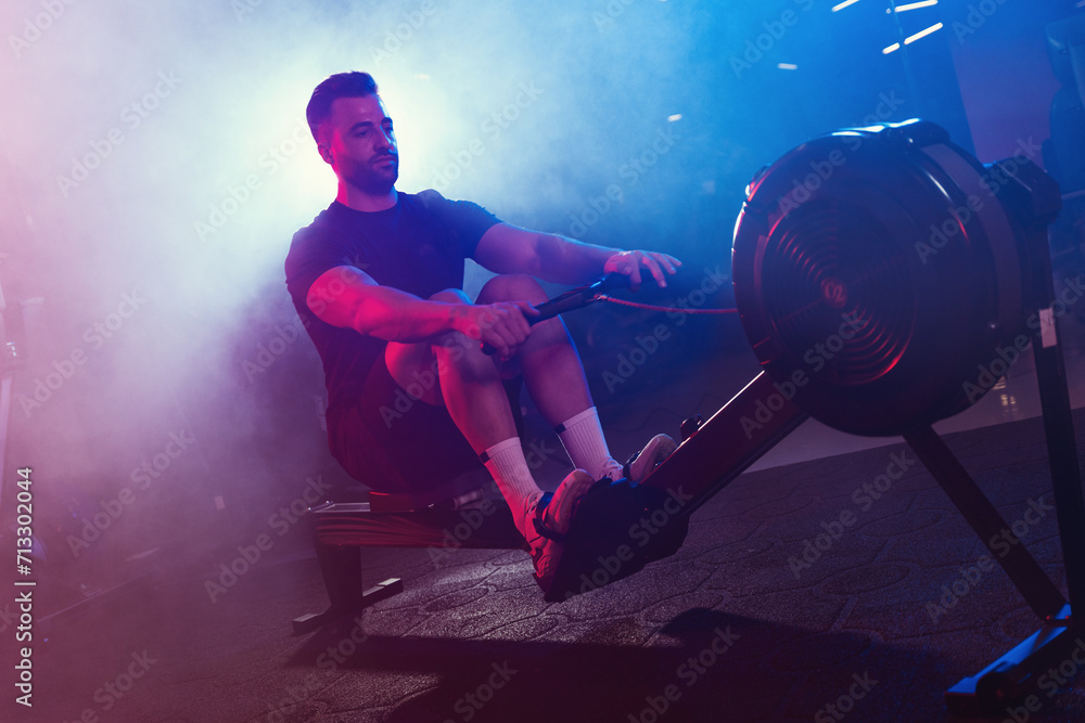 A male athlete pulls the oars on a rowing machine, his form lit by ...