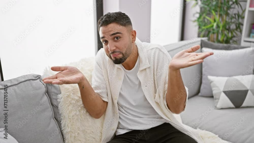 Baffled young arab man indoors, sitting cluelessly on living room sofa ...