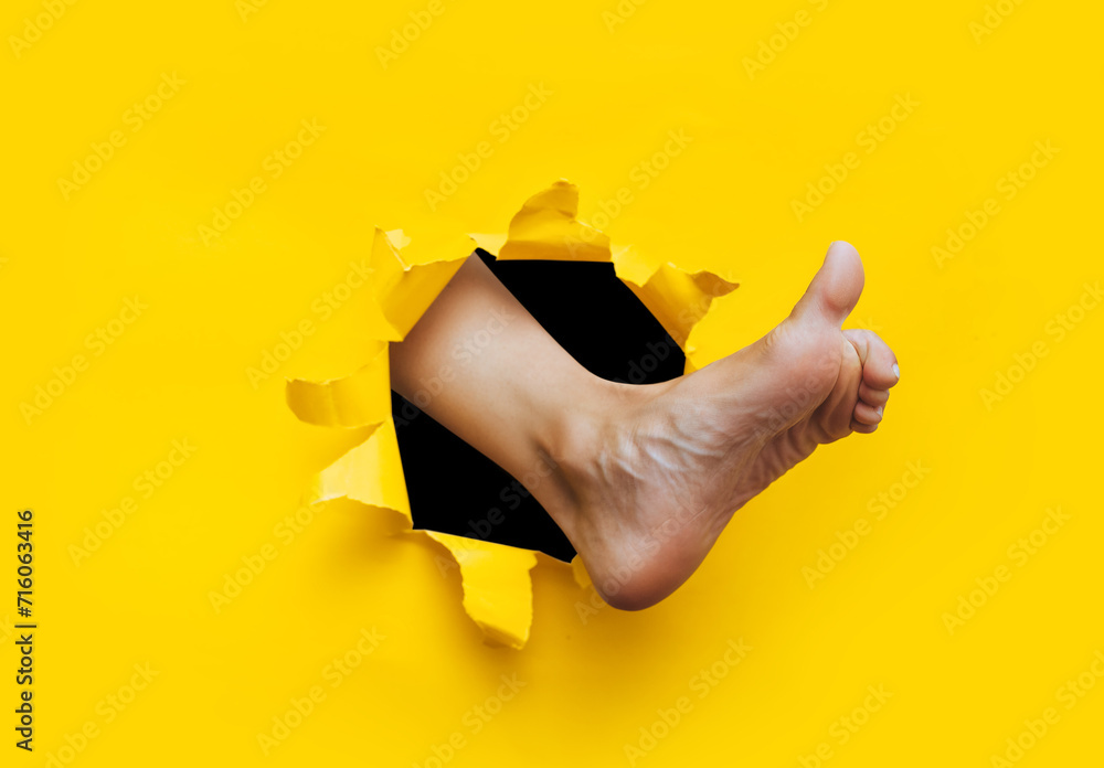 A left woman's foot appears in a torn hole in yellow paper and tries to ...