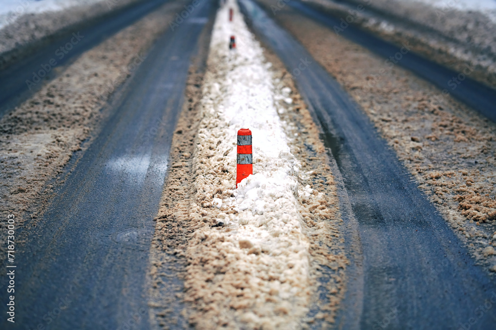 Asphalt roadway covered with slushy snow and orange plastic pole with ...