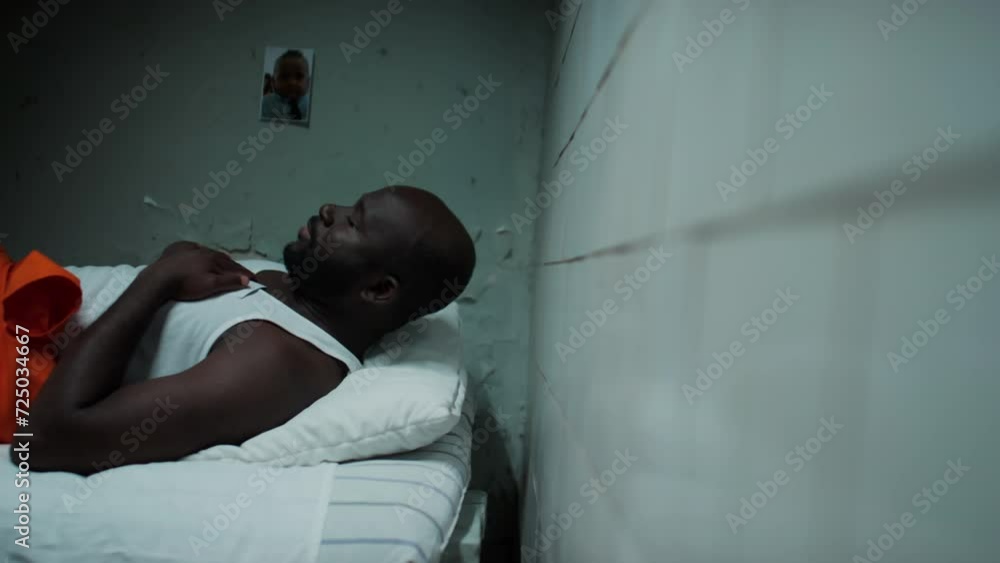 Unhappy Black inmate lying on bed in prison, looking at photo of child ...