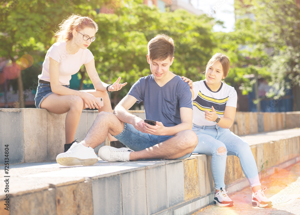 Interested teen boy sitting with phone on summer city street with two ...