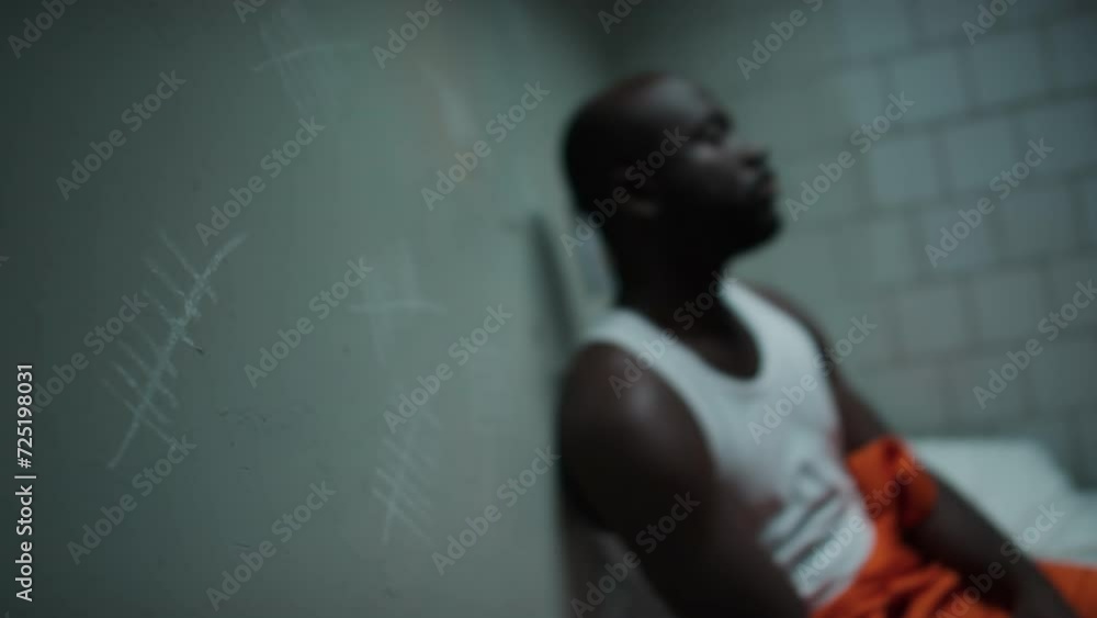 Rack focus shot of tired Black inmate sitting on bed in prison cell and ...