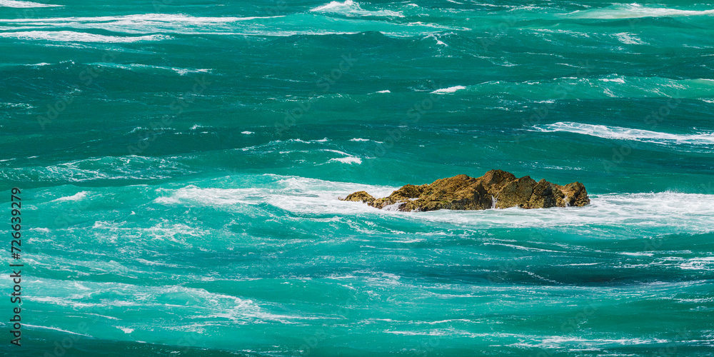 Turquoise ocean with rough water, waves and a rock protruding above the ...