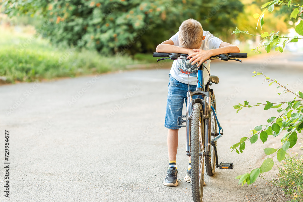Tired boy in a t-shirt and denim shorts leans on his bicycle after a ...