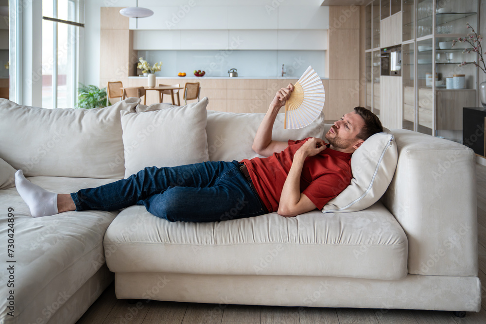 Overheated perspiring man lying on sofa, waving with hand fan ...