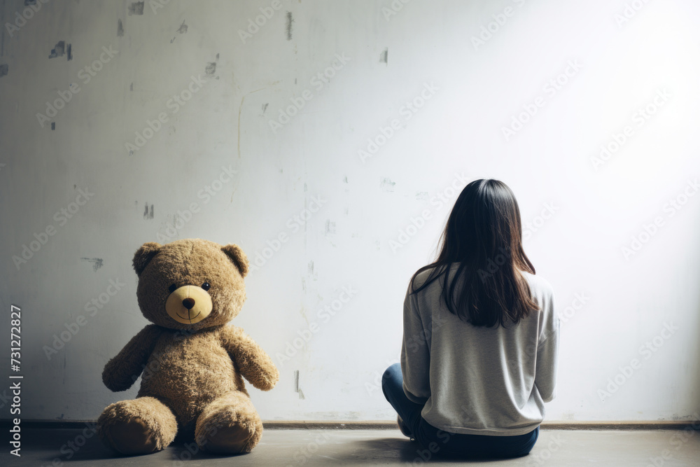 girl behind body with teddy bear sitting and facing an empty white wall ...