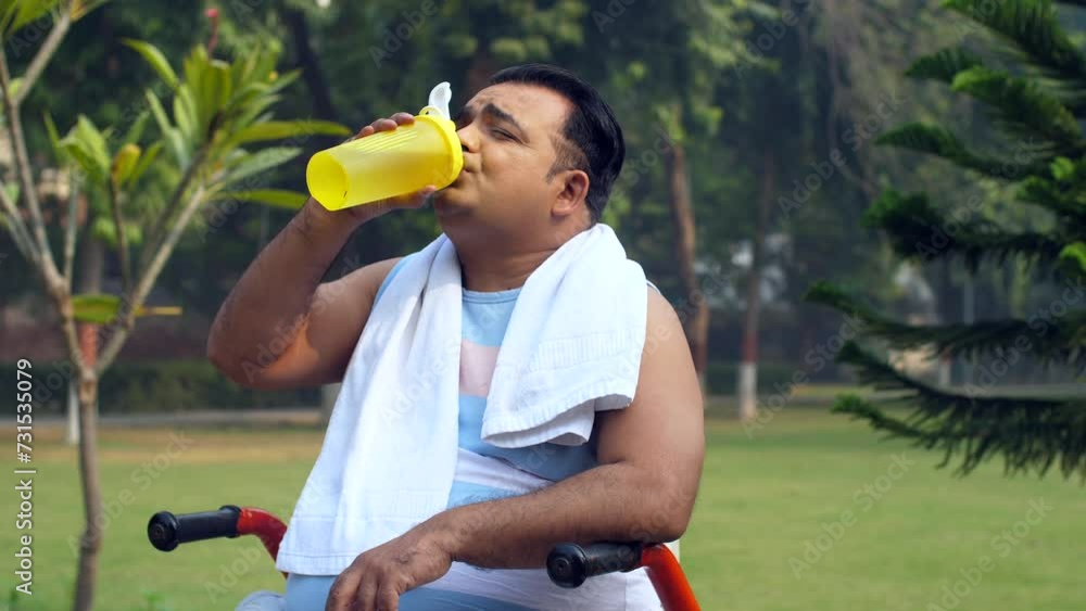 Portrait of an obese indian man sitting in a park and drinking water ...