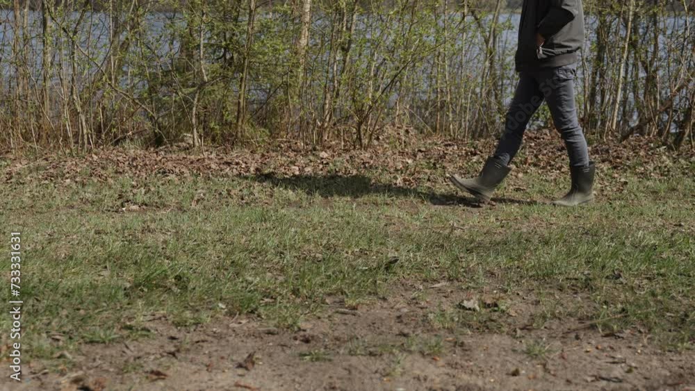 Unrecognizable slender man in rubber boots walks along bushes on shore ...