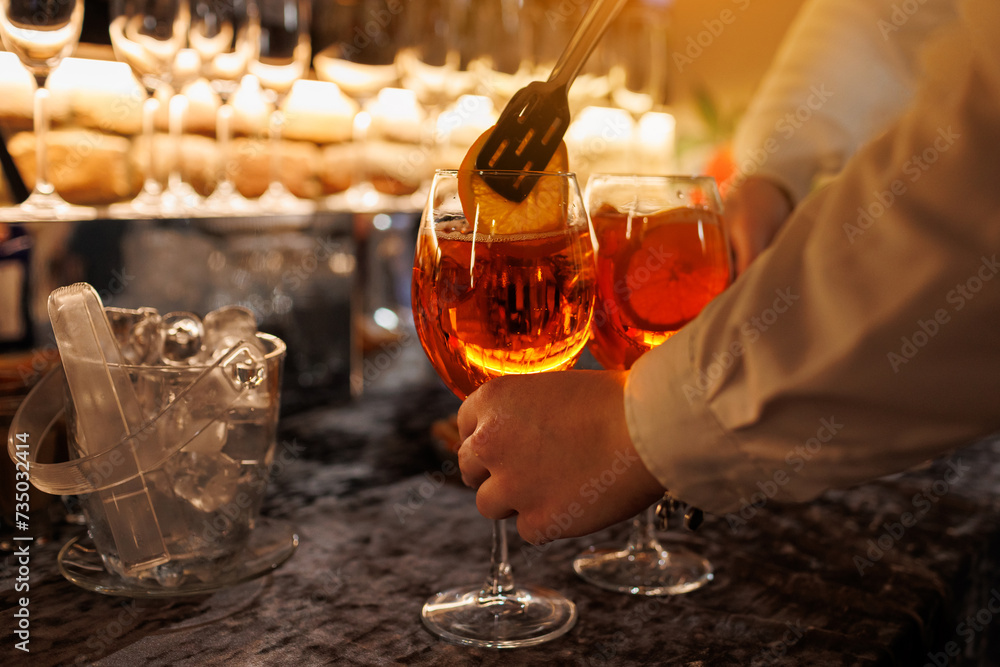Bartender makes two glasses of cocktail Aperol spritz on bar counter, adds fresh orange slices ...