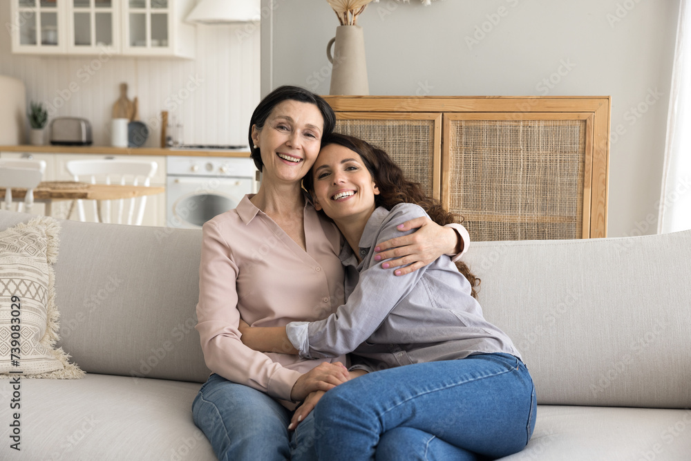 Happy beautiful grown-up daughter cuddling elderly mother laugh sit together on sofa indoors ...