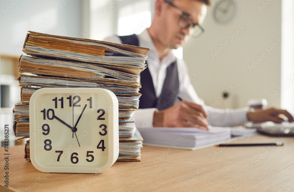 Stack of papers, table clock and businessman engrossed in work ...