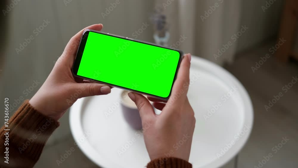 Young woman sitting at home holding smartphone green mock-up screen in ...