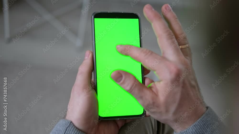Young man sitting at home holding smartphone green mock-up screen in ...