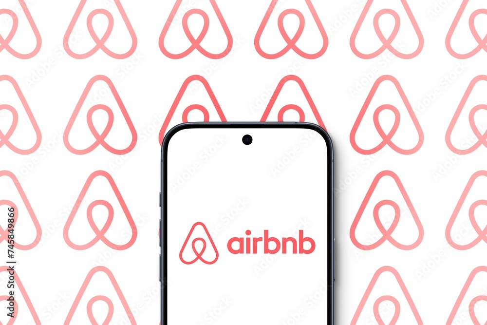 airbnb Logo is displayed on a modern smartphone, many pink Airbnb Logos in front of a white ...