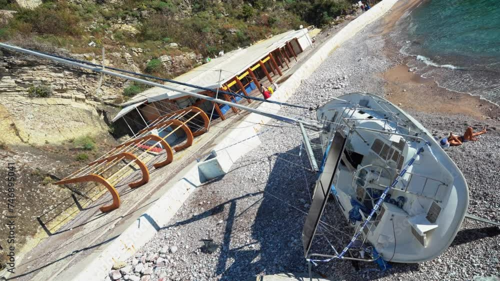 Carefree tourist sunbathes next to beached sunken yacht on pebble beach in off-season near ...