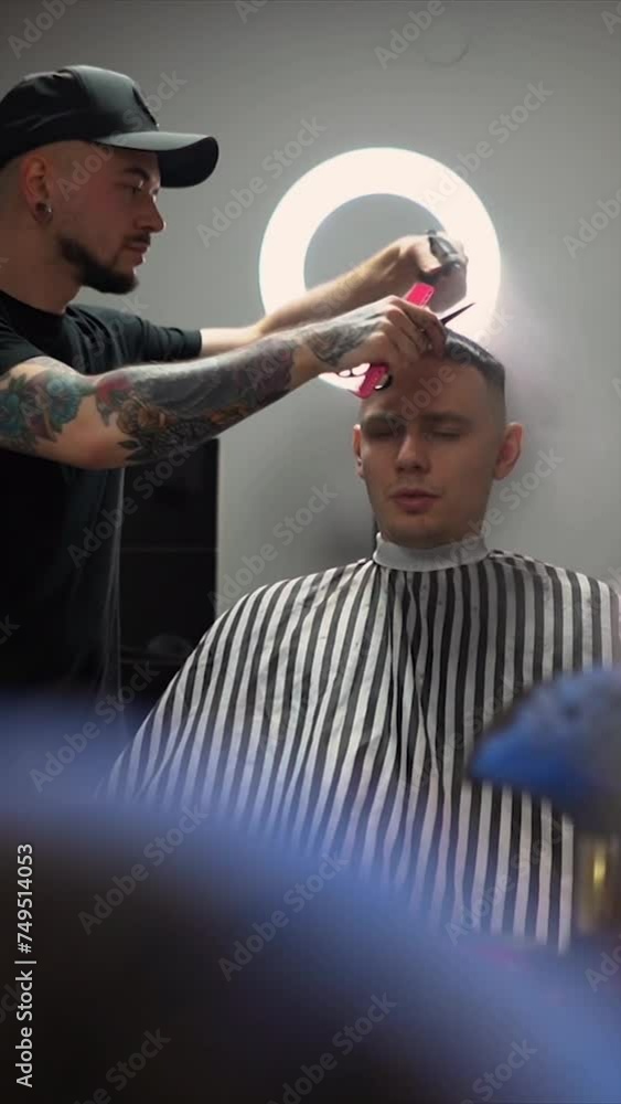 A barber man with a tattoo on his arm makes a haircut to a brunette guy ...