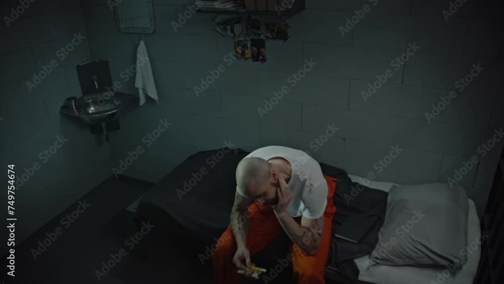 Prisoner in orange uniform sits on bed in prison cell and looks on ...