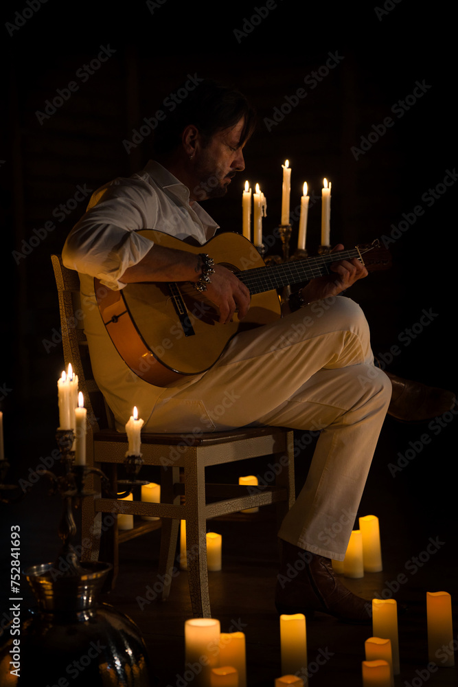 Rear view of a man dressed in white playing the Spanish guitar at home by candlelight, learning ...