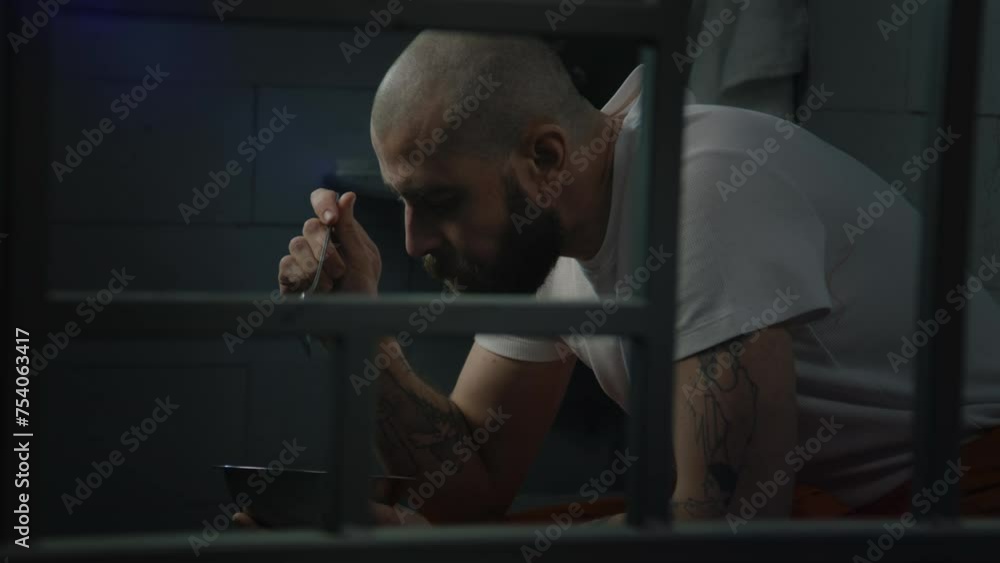 Inmate in tattoos in orange uniform sits on bed in jail cell, eats ...