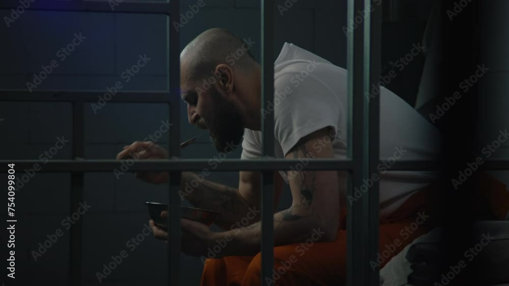 Male prisoner in orange uniform sits on bed in prison cell, eats ...