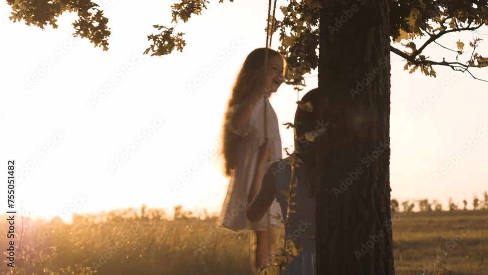 Mother gently pushes daughter on swing standing behind massive tree in ...