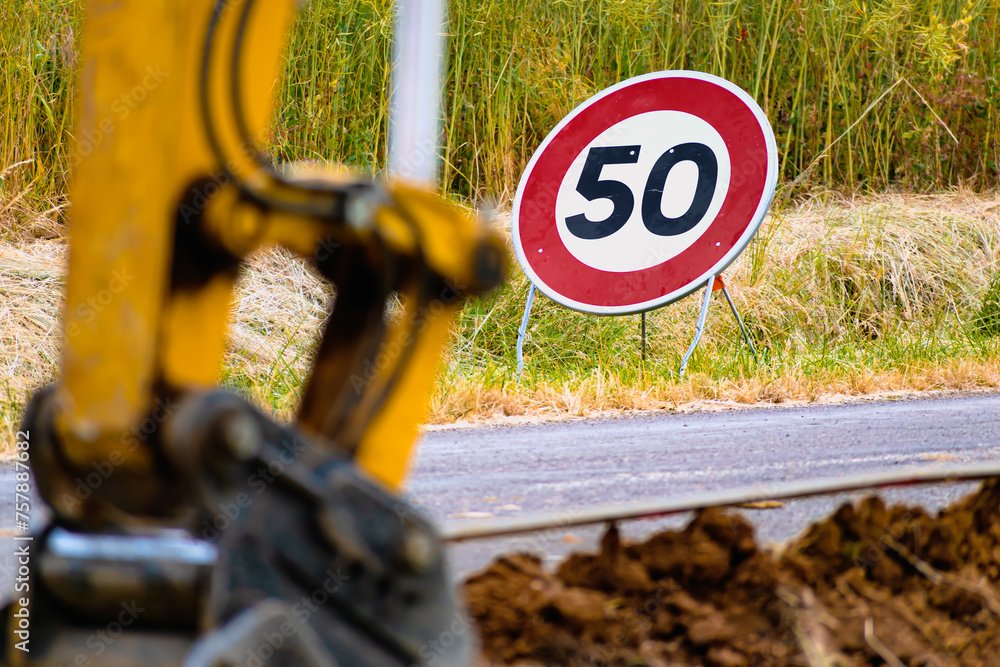 Arm of a mini digger and bucket with a speed limit sign at 50, road ...