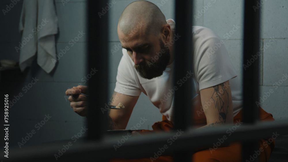Inmate in tattoos in orange uniform sits on bed in jail cell, eats ...