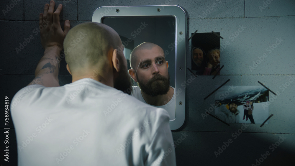 Depressed male prisoner stands in prison cell, looks at the pictures ...