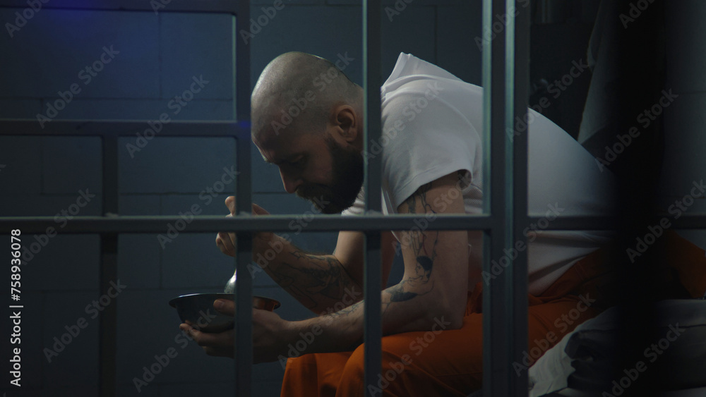 Male prisoner in orange uniform sits on bed in prison cell, eats ...