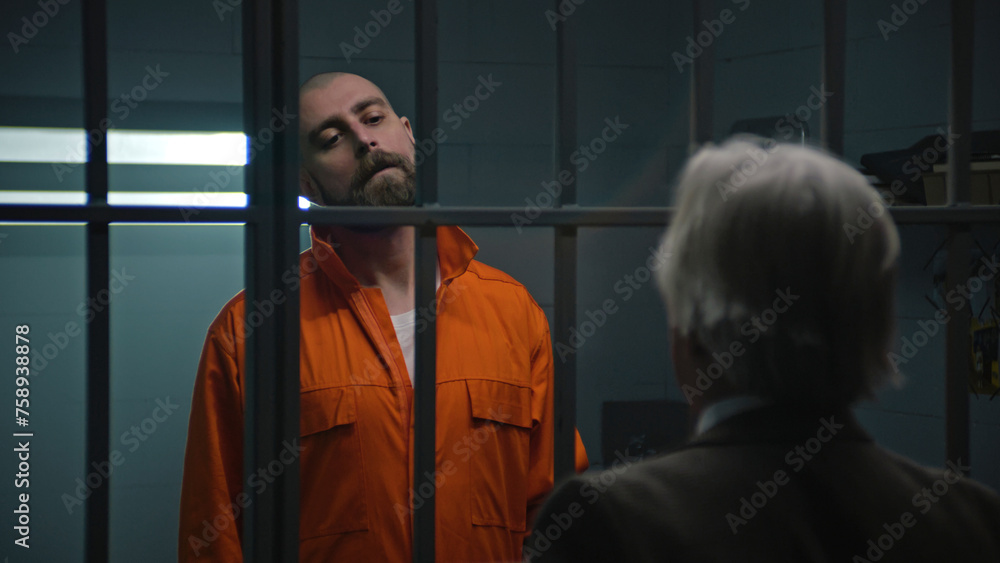 Gangster in orange uniform stands behind bars in prison cell, talks ...