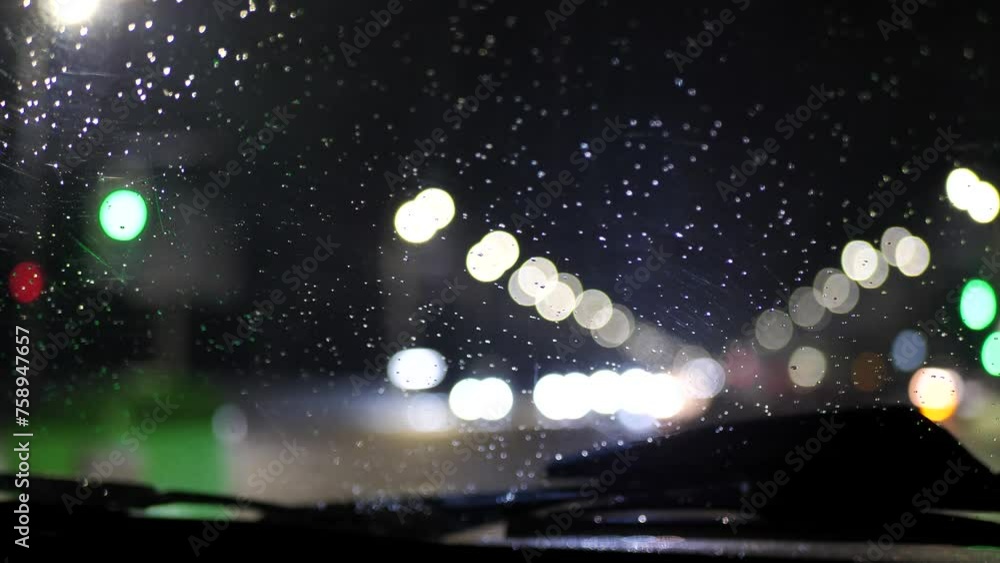 Night city through the wet window of a moving car with a wiper. Rainy ...