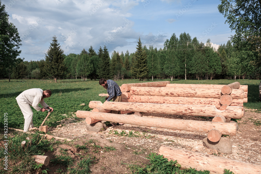Two men are building a wooden house from logs in nature, cutting logs ...
