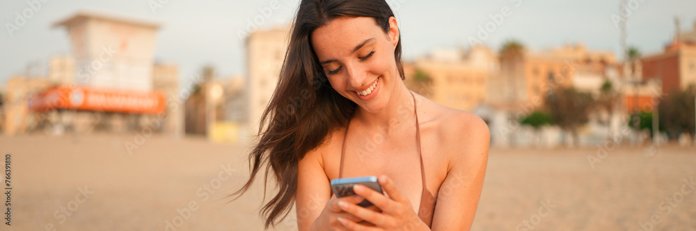 Slim beautiful brown-haired woman with long hair is using mobile phone sitting on the beach ...