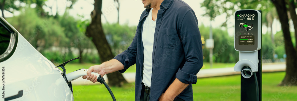 Man recharge EV electric vehicle's battery from EV charging station in outdoor green city park ...