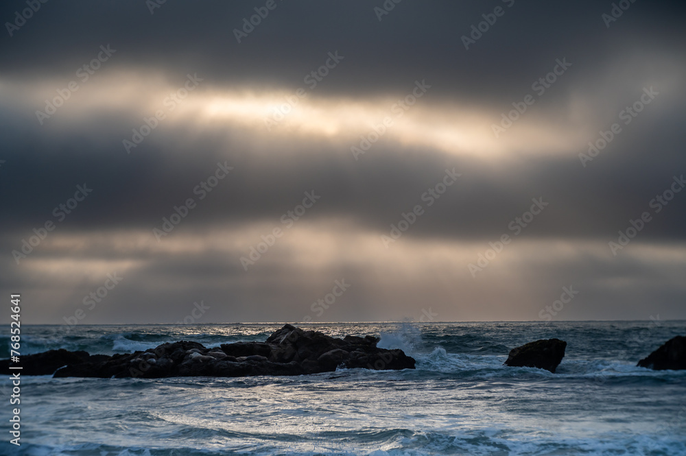 Sun rays coming through gray clouds over ocean waves and coastal rocks ...