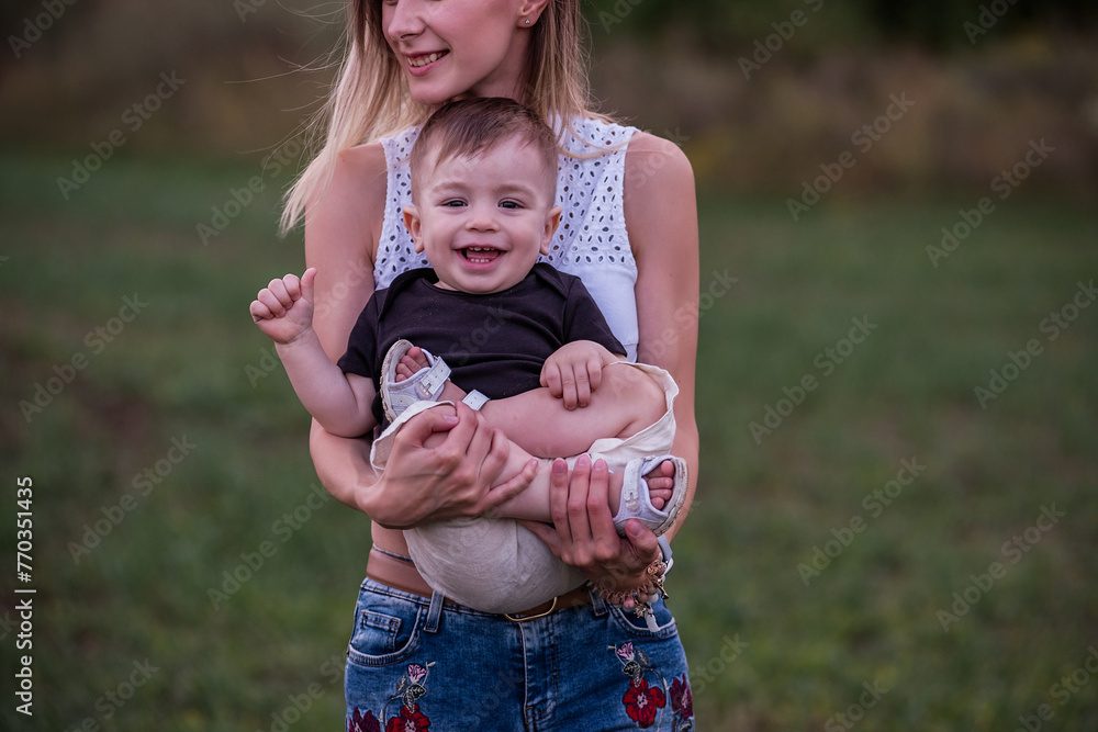 Young woman smiles, holds toddler son in her arms, standing in lush ...