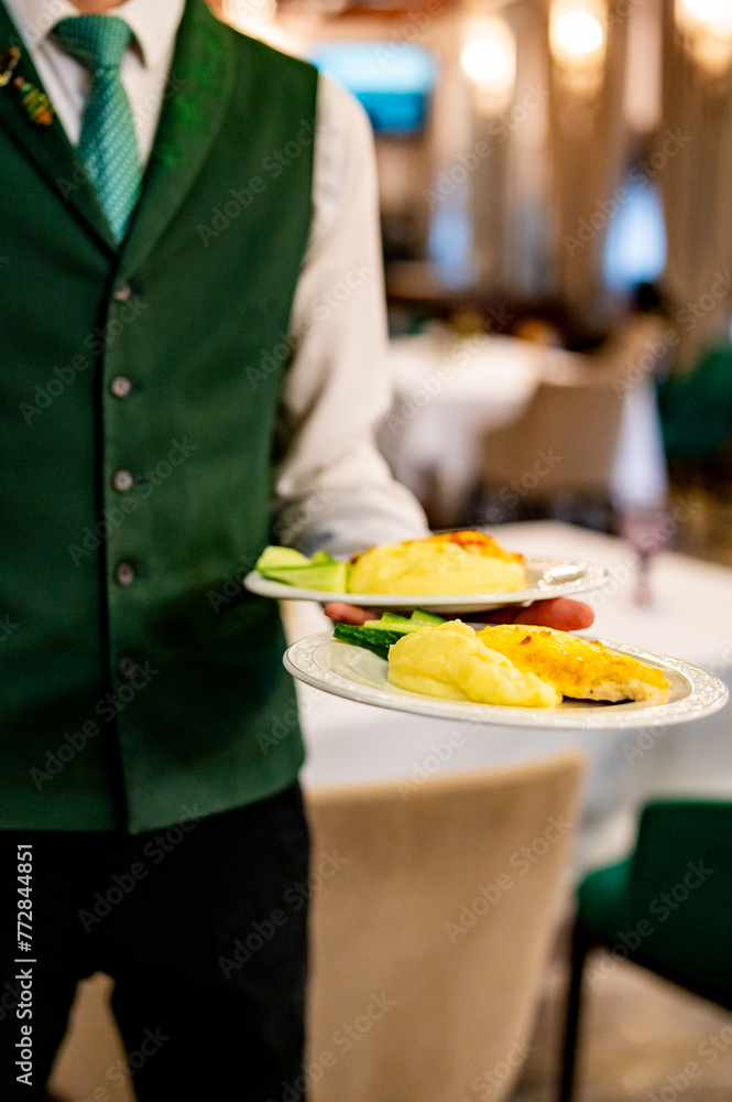 A waiter in a green vest and tie serves two plates of delicious food in ...